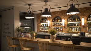 Uma área de bar com cadeiras de madeira, parede de tijolos e prateleiras com garrafas.