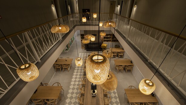 Uma ampla sala de jantar com pé direito alto, várias mesas de madeira e luminárias de tecido penduradas.