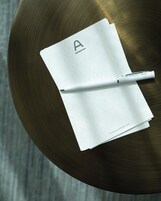 Bloco de notas branco com padrão geométrico e uma caneta sobre uma mesa redonda de madeira escura.