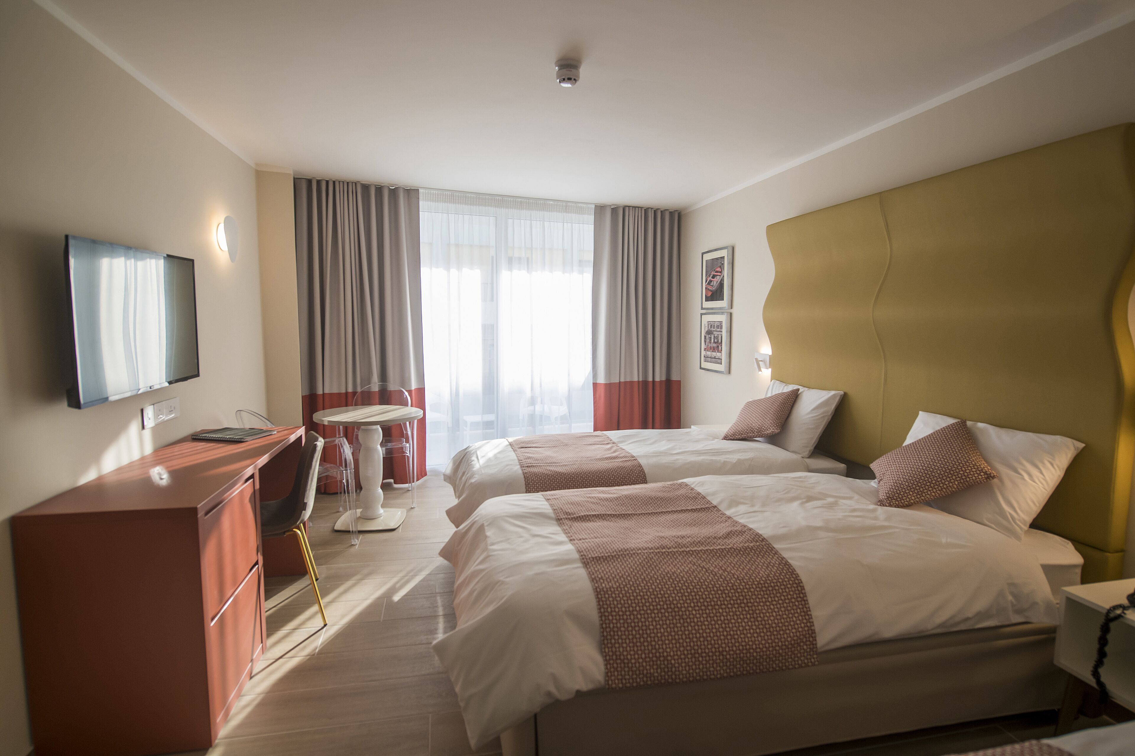 Quarto de hotel com duas camas, uma escrivaninha, uma cadeira, uma televisão e uma janela com cortinas.
