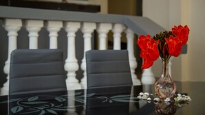 Uma mesa de jantar com um vaso de flores vermelhas, cadeiras pretas e um parapeito branco ao fundo.
