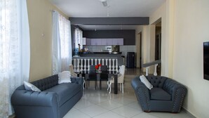 Uma sala de estar com sofá, poltronas, uma mesa de jantar e uma cozinha ao fundo.