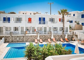 Área da piscina com piscina azul cristalina, edifícios brancos com persianas azuis e vermelhas, e uma parede de pedra.