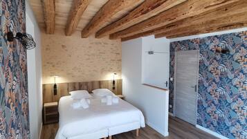 Quarto com teto de madeira, cama com roupa de cama branca, mesa de cabeceira, luminária na parede e papel de parede estampado em uma parede.