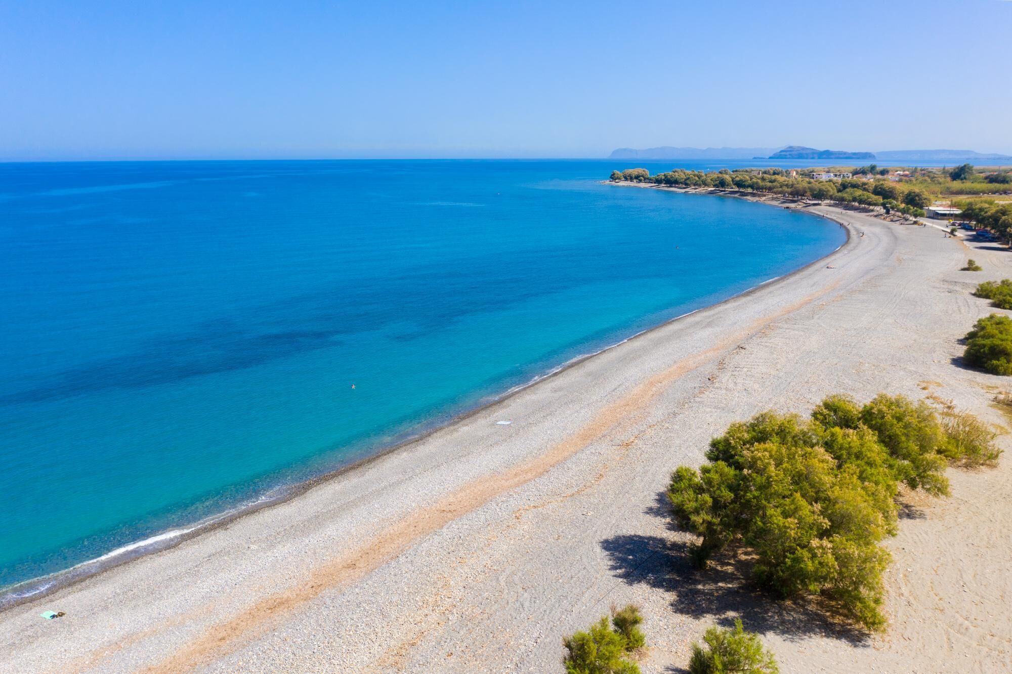Uma praia costeira com águas azul-claras, areal arenoso e vegetação rasteira.