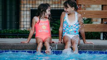 Duas crianças sentadas à beira da piscina, uma de fato de banho e a outra de vestido.
