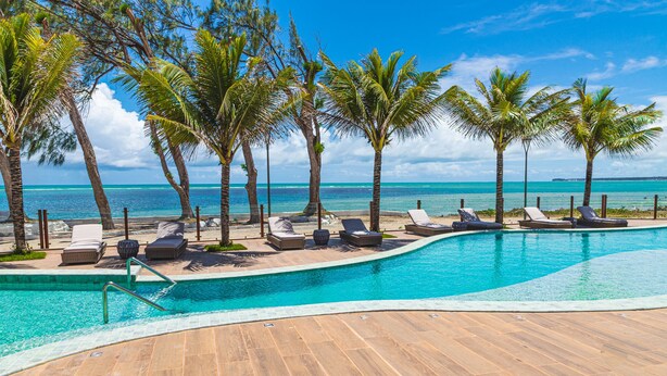 Uma área à beira da piscina com espreguiçadeiras e palmeiras, com vista para um oceano azul e límpido.