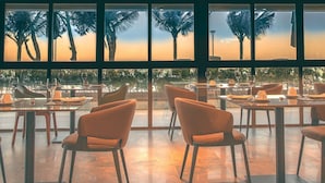 Restaurante com grandes janelas que oferecem vista para palmeiras e um pôr do sol. Mesas montadas com cadeiras e louças.