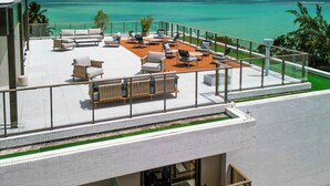 Terraço no telhado com área de estar externa, vista para o oceano e guarda-corpo de vidro.