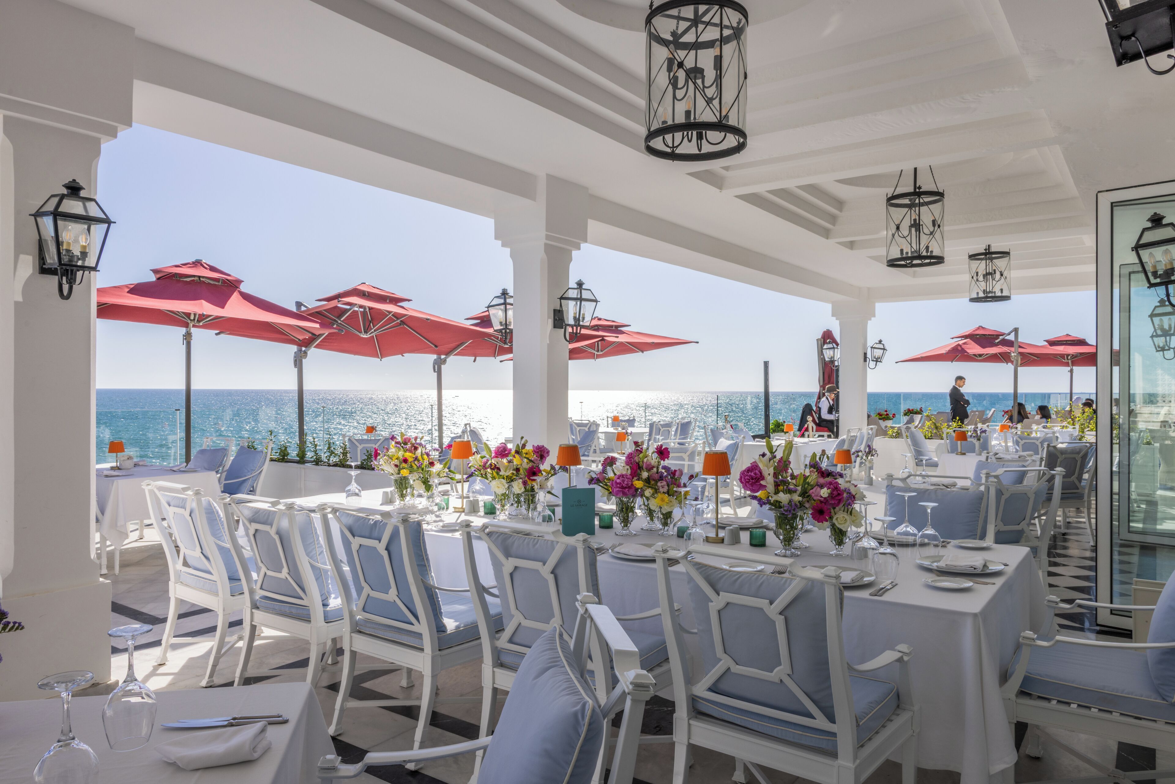 Restaurante à beira-mar com mesas externas, toalhas brancas e guarda-sóis vermelhos.