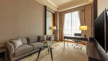 Quarto de hotel com sofá, mesa de centro de vidro, escrivaninha com cadeira, televisão e janela com cortinas.