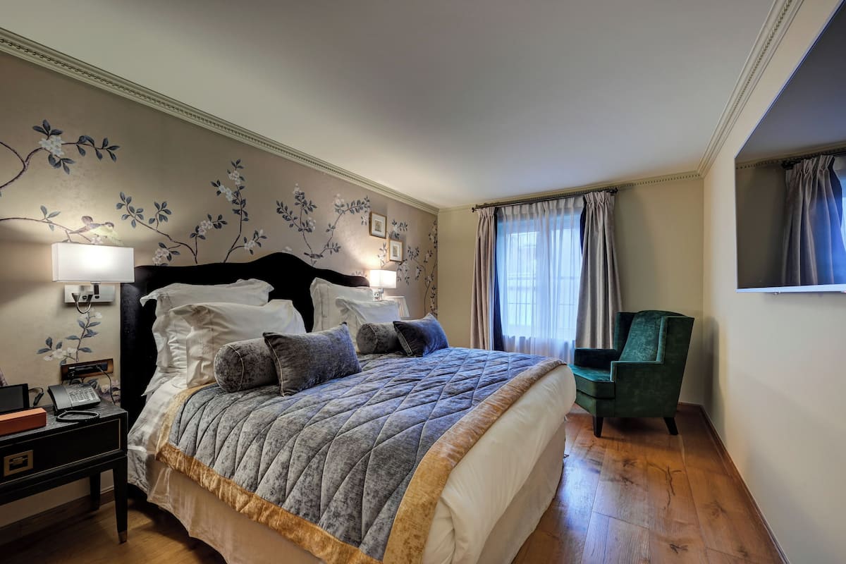 Um quarto bem arrumado com uma cama grande, uma mesa de cabeceira com abajur, uma poltrona verde e uma janela com cortinas.