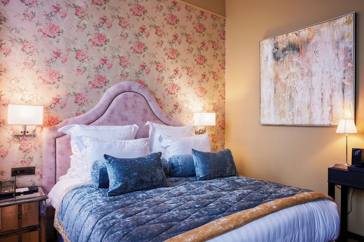 Quarto com papel de parede floral, cabeceira rosa, colcha acolchoada azul e duas luminárias de cabeceira.