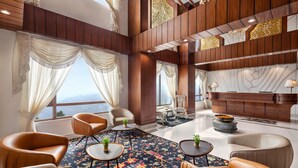 Um hall espaçoso com teto alto, painéis de madeira e grandes janelas com vista para as montanhas. Há vários espaços de estar com poltronas de couro e pequenas mesas, e um balcão de recepção com design moderno.