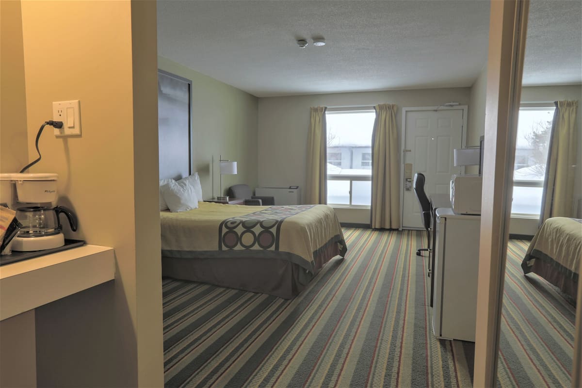 Quarto de hotel com cama, escrivaninha, cafeteira, geladeira e janela com cortinas.
