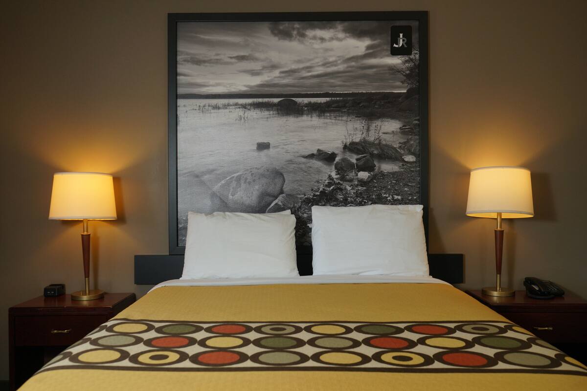 Quarto de hotel com cama, duas luminárias de cabeceira, mesa de cabeceira com telefone e uma moldura grande na parede.