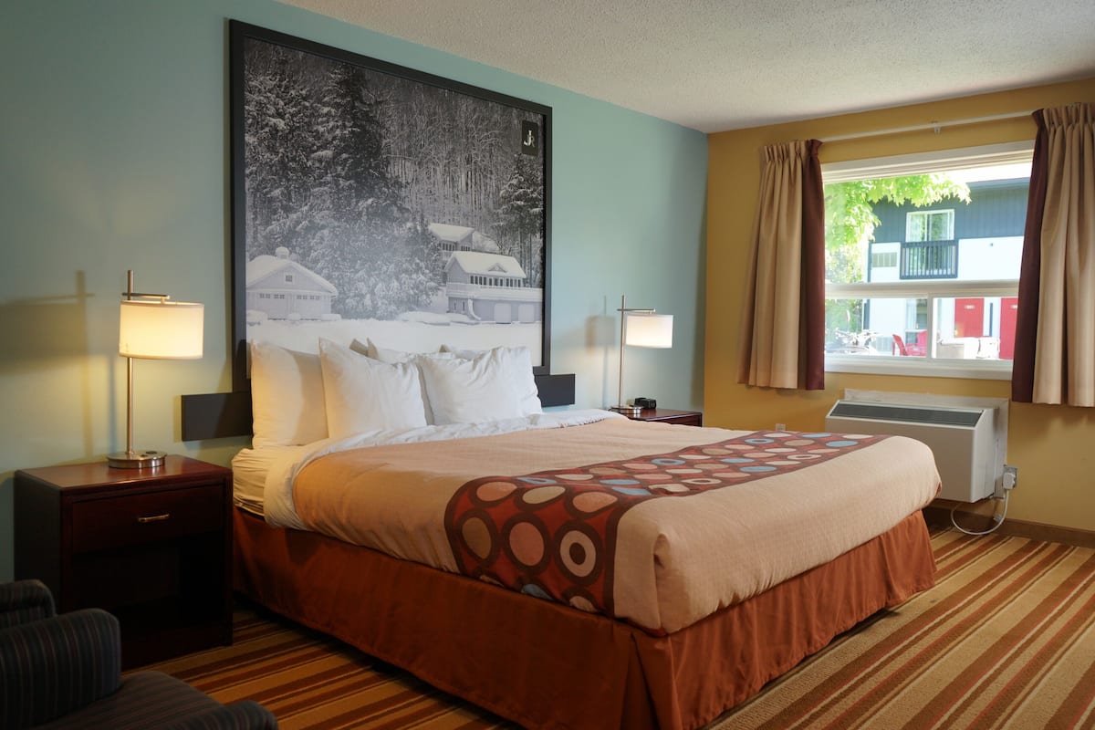 Quarto de hotel com uma cama grande, duas mesas de cabeceira, um criado-mudo, uma janela com cortinas e um ar condicionado fixado na parede.