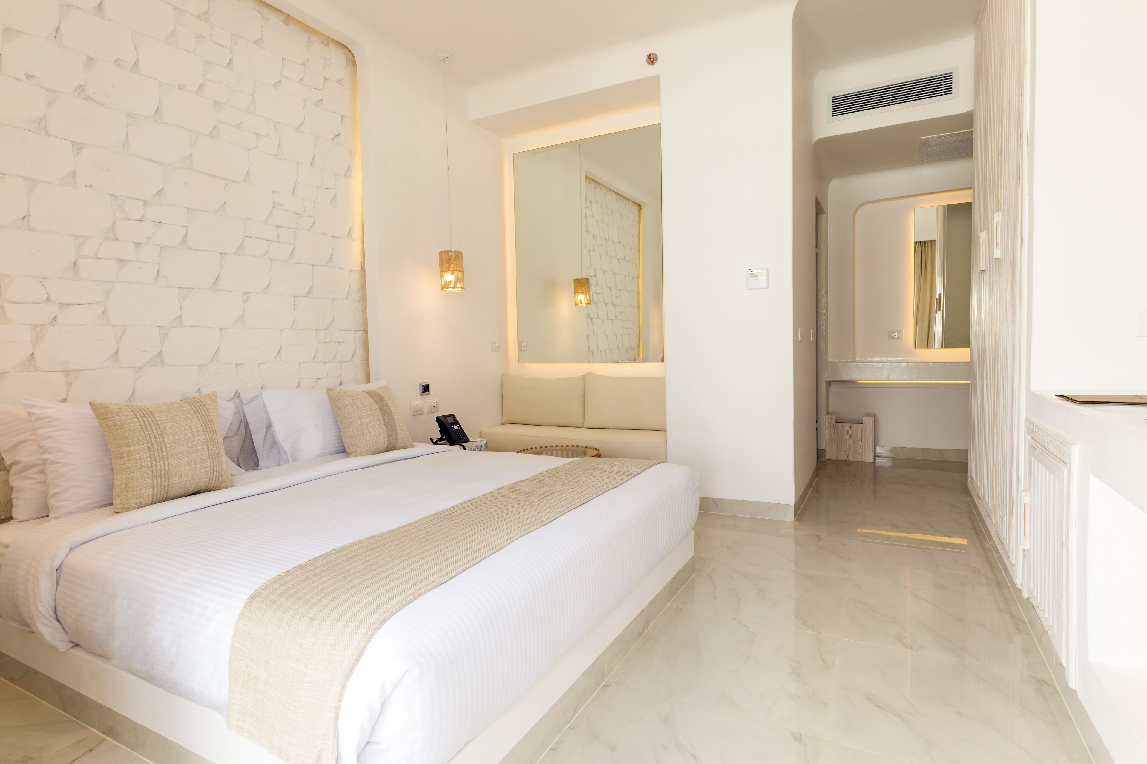 Quarto de hotel moderno com uma cama grande, área de estar, espelho e banheiro com chuveiro.