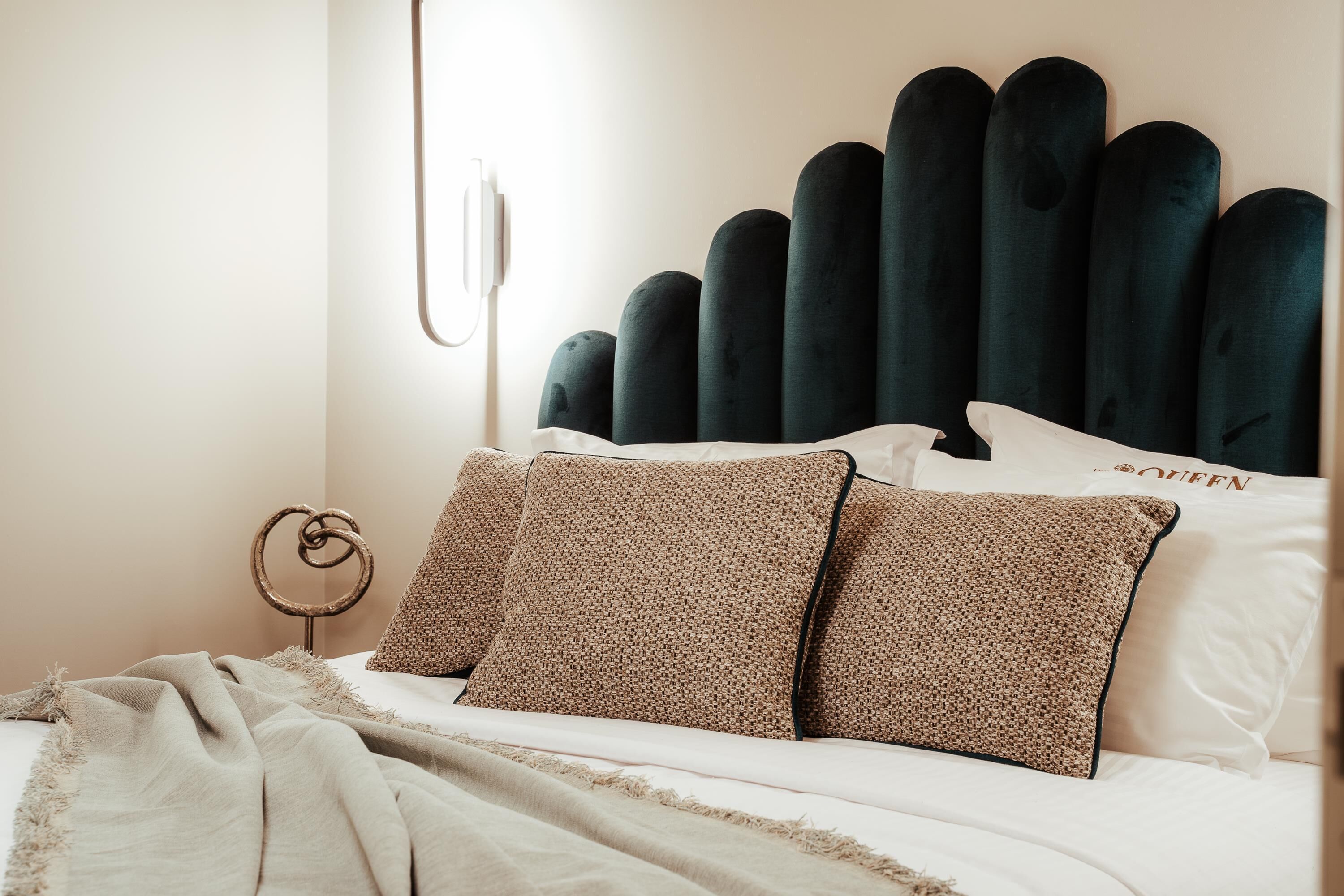 Uma cama com cabeceira escura, almofadas bege e um cobertor branco.