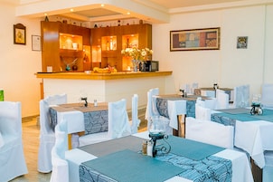 Um restaurante com várias mesas, cada uma coberta por toalhas brancas e jogos americanos azuis. Há uma área de bar com balcão de madeira e diversos itens expostos.