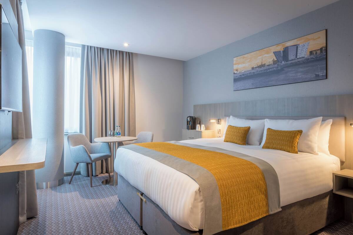 Quarto de hotel com uma cama grande, uma escrivaninha, uma cadeira e uma mesinha.