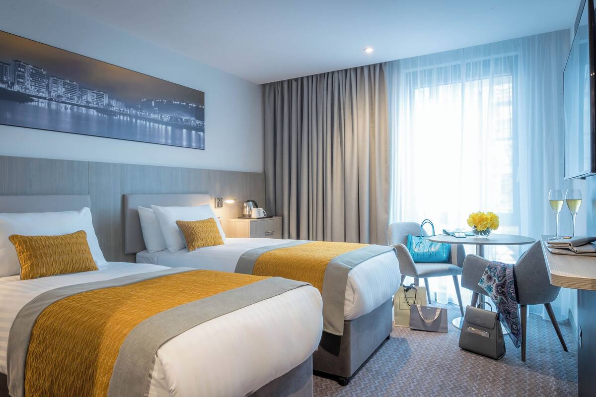 Quarto de hotel com duas camas, uma escrivaninha, uma cadeira e vista para a paisagem urbana.
