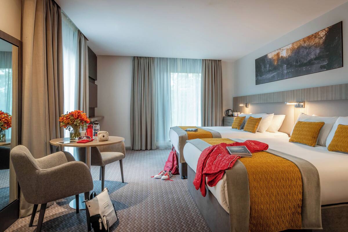 Quarto de hotel com uma cama grande, duas mesas de cabeceira, um espaço de estar com uma poltrona, uma mesinha com um vaso de flores e um quadro na parede.