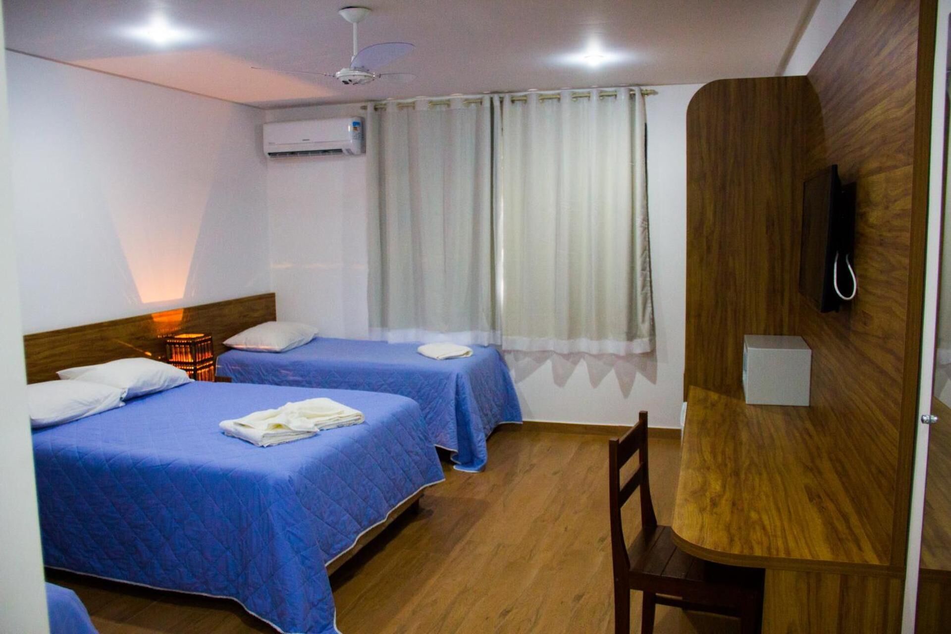Quarto de hotel com duas camas, uma escrivaninha de madeira, uma cadeira, uma televisão fixada na parede e um aparelho de ar condicionado.