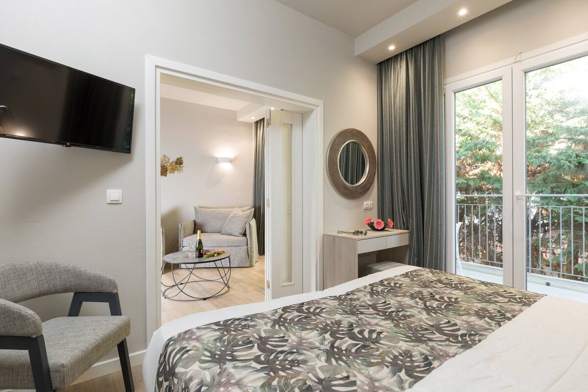 Quarto de hotel moderno com uma cama grande, televisão na parede, varanda com vista para árvores e um pequeno espaço de estar com mesa e cadeira.