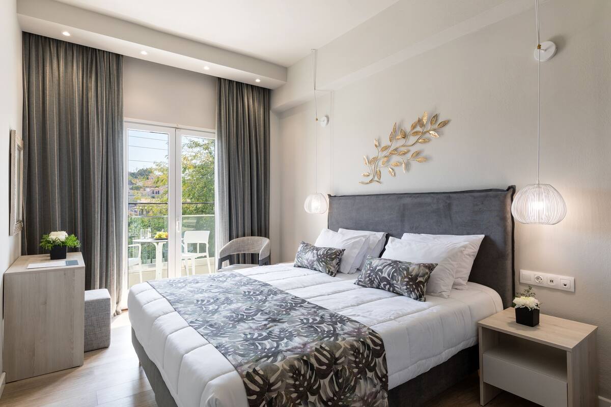 Quarto moderno com uma cama grande, uma mesinha de cabeceira, uma cadeira e vista para o exterior.