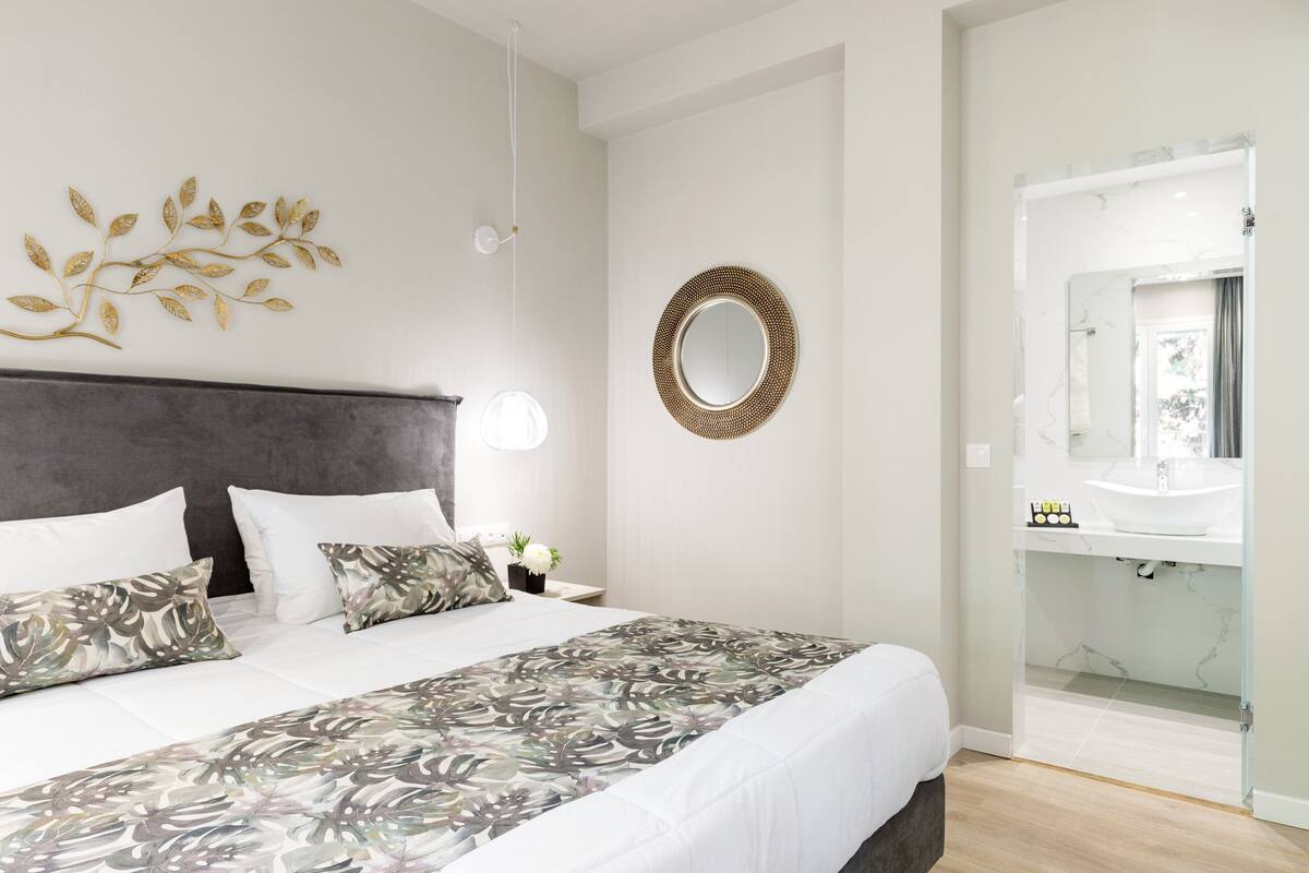 Quarto moderno com cama grande, mesa de cabeceira com luminária, um objeto decorativo na parede e banheiro com pia e espelho.