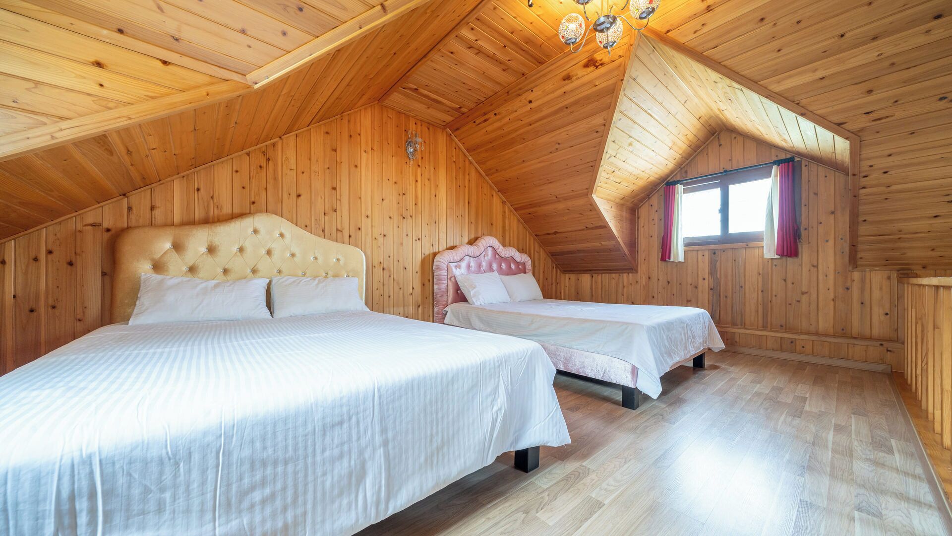 Quarto com duas camas, paredes de madeira, teto inclinado e janela com cortinas.