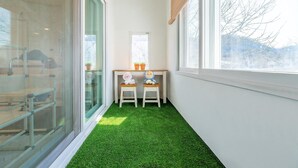 Quarto com porta de vidro, uma pequena escrivaninha com duas cadeiras e janela com vista para árvores.
