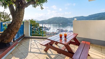 Terraço com mesa e bancos de madeira, com vista para um corpo d'água e colinas.
