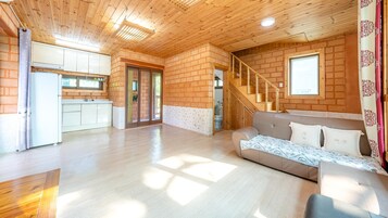 Uma sala de estar ampla com teto de madeira, uma cozinha com armários brancos, um sofá com almofadas estampadas e uma escada que leva a um andar superior.