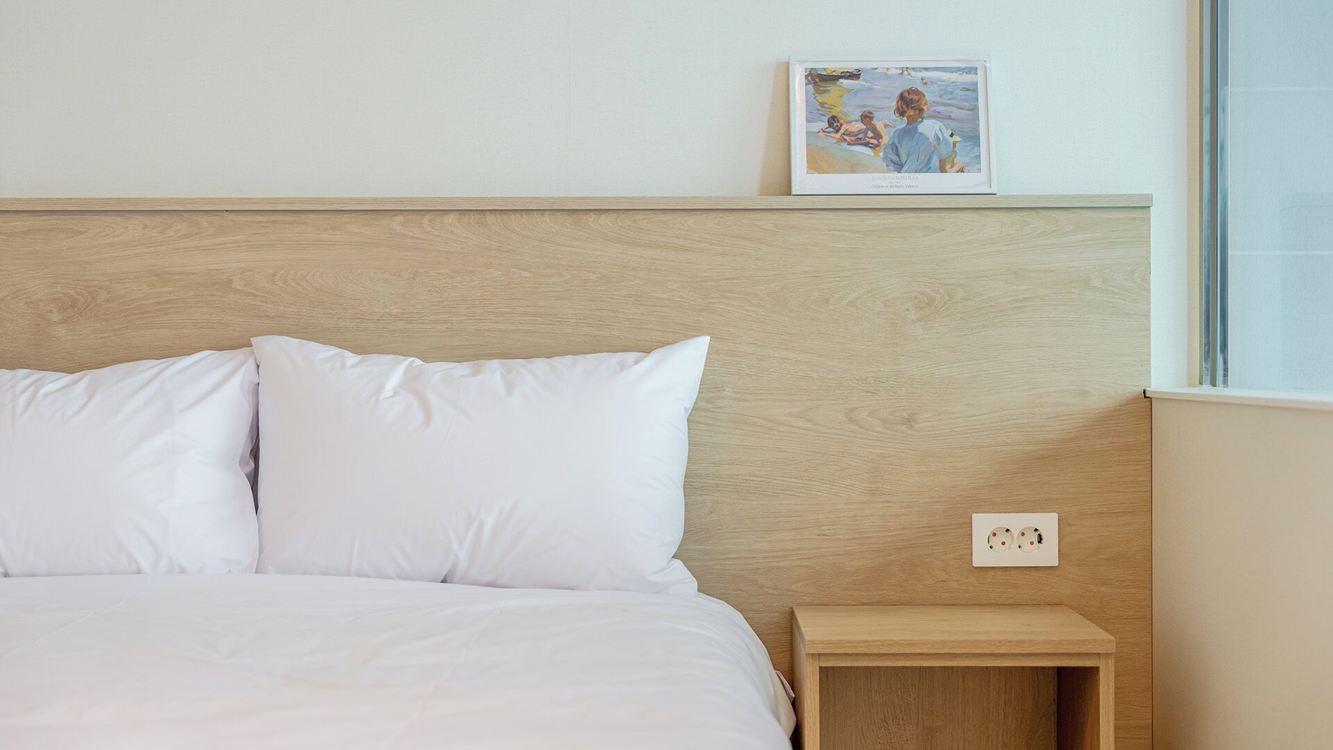 Uma cama bem arrumada com lençóis brancos, cabeceira de madeira e uma pequena mesa de cabeceira com tomada.