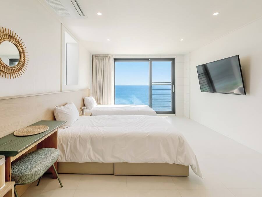 Quarto de hotel com cama, escrivaninha, cadeira, televisão e uma janela grande com vista para o mar.