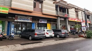 Um edifício com várias lojas, incluindo uma farmácia e uma loja de artigos gerais. Vários carros estão estacionados na frente, e uma motocicleta é visível na calçada.