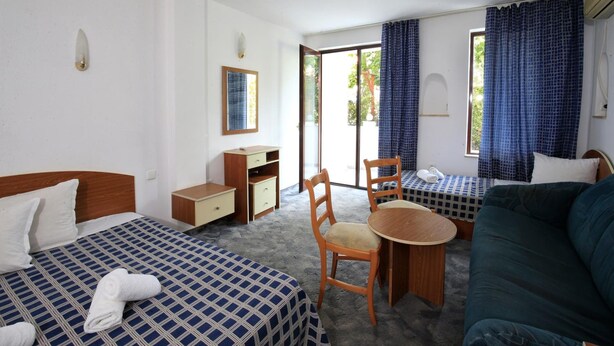 Quarto de hotel com duas camas, um sofá, uma mesa pequena e uma cadeira. Há uma janela com cortinas, um espelho e uma mesa de cabeceira.