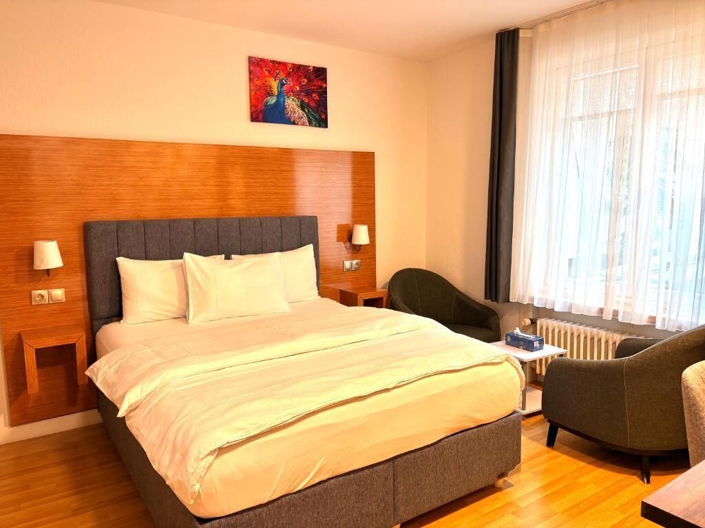 Quarto de hotel com uma cama grande, cabeceira de madeira, um quadro acima da cama, uma cadeira, uma mesa e uma janela com cortinas.