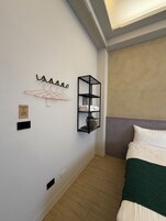 Quarto de hotel com uma cama, prateleira fixada na parede com toalhas dobradas, tomada e um aviso com informações sobre o Wi-Fi.