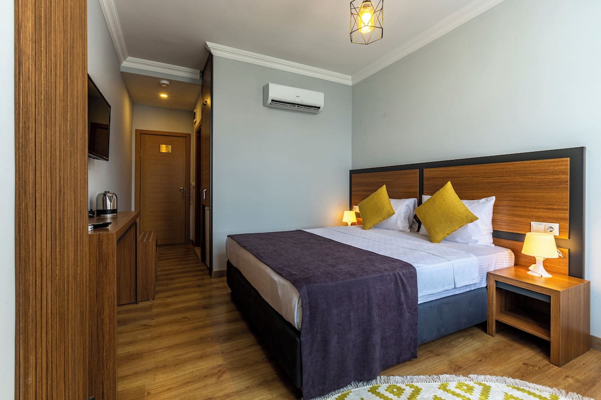 Quarto de hotel com piso de madeira, cama com cabeceira escura, mesa de cabeceira com abajur e aparelho de ar condicionado na parede.