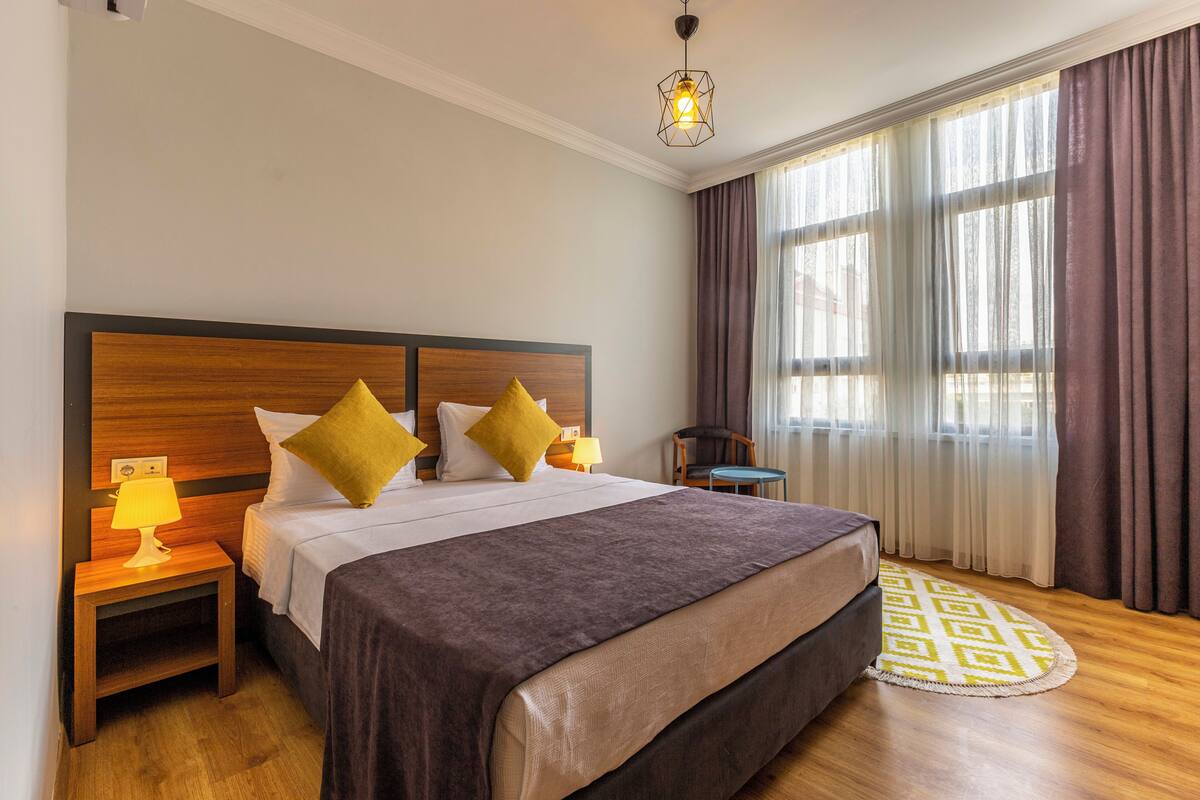 Quarto de hotel com uma cama grande, duas mesas de cabeceira com luminárias, uma cabeceira de madeira, uma pequena mesa e uma janela com cortinas.