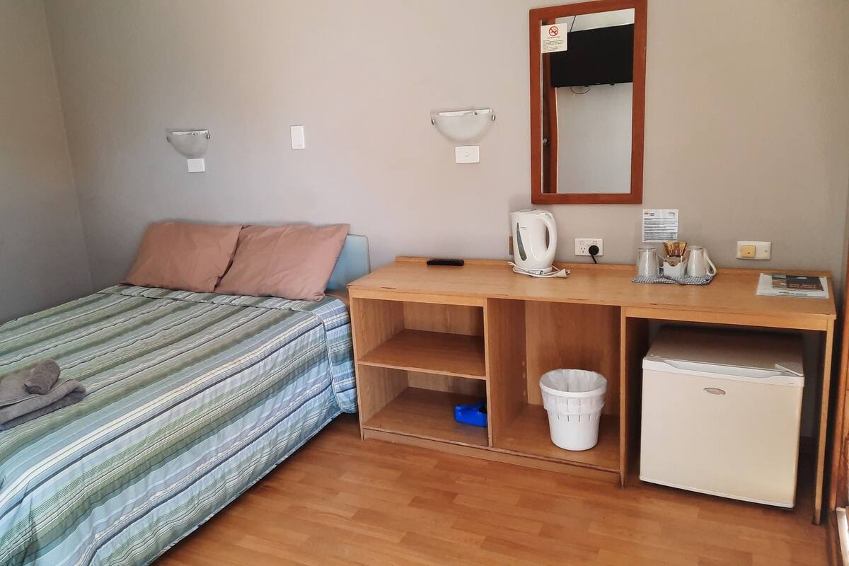 Quarto pequeno e arrumado, com uma cama, uma escrivaninha de madeira, um espelho, uma chaleira e um refrigerador compacto.