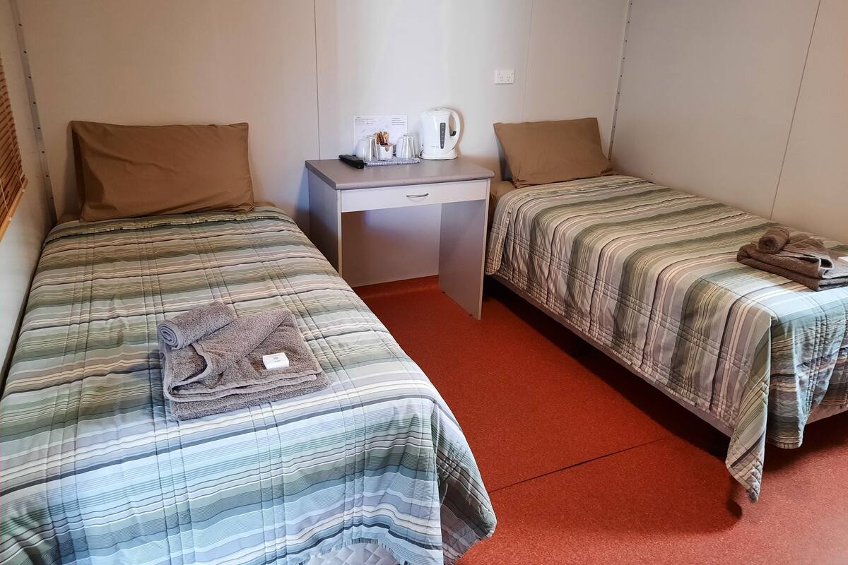 Quarto com duas camas, cada uma com roupa de cama listrada, uma escrivaninha com uma chaleira e um controle remoto, e um par de toalhas dobradas em uma das camas.