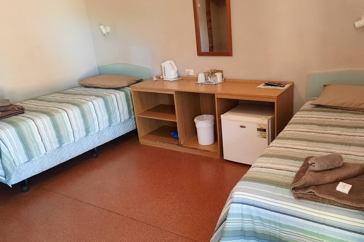 Quarto com duas camas, uma escrivaninha pequena, um espelho e uma lixeira.