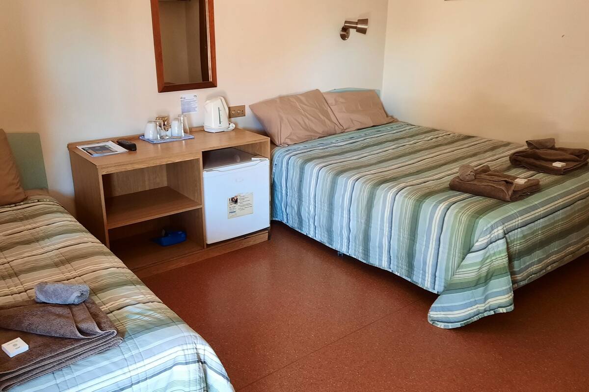 Quarto pequeno e compacto com duas camas de solteiro, uma escrivaninha de madeira com geladeira, um espelho e uma pequena prateleira.