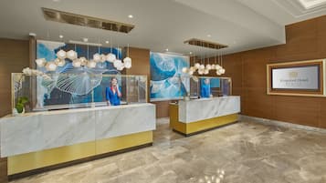 Uma área de recepção de hotel com balcões de mármore, uma grande placa emoldurada e um lustre moderno.