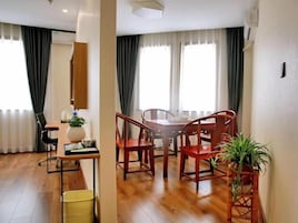 Quarto com mesa de jantar de madeira e cadeiras, uma pequena escrivaninha com cadeira, uma planta em vaso e uma lixeira.