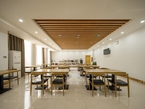 Uma sala de conferências moderna com mesas e cadeiras de madeira, uma TV de tela plana na parede e uma área de cozinha ao fundo.